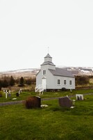 Uma igreja branca com um campanário, rodeada por um campo gramado com lápides e uma montanha ao fundo.