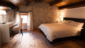 Quarto com parede de pedra, vigas de madeira no teto, cama, escrivaninha e cadeira.