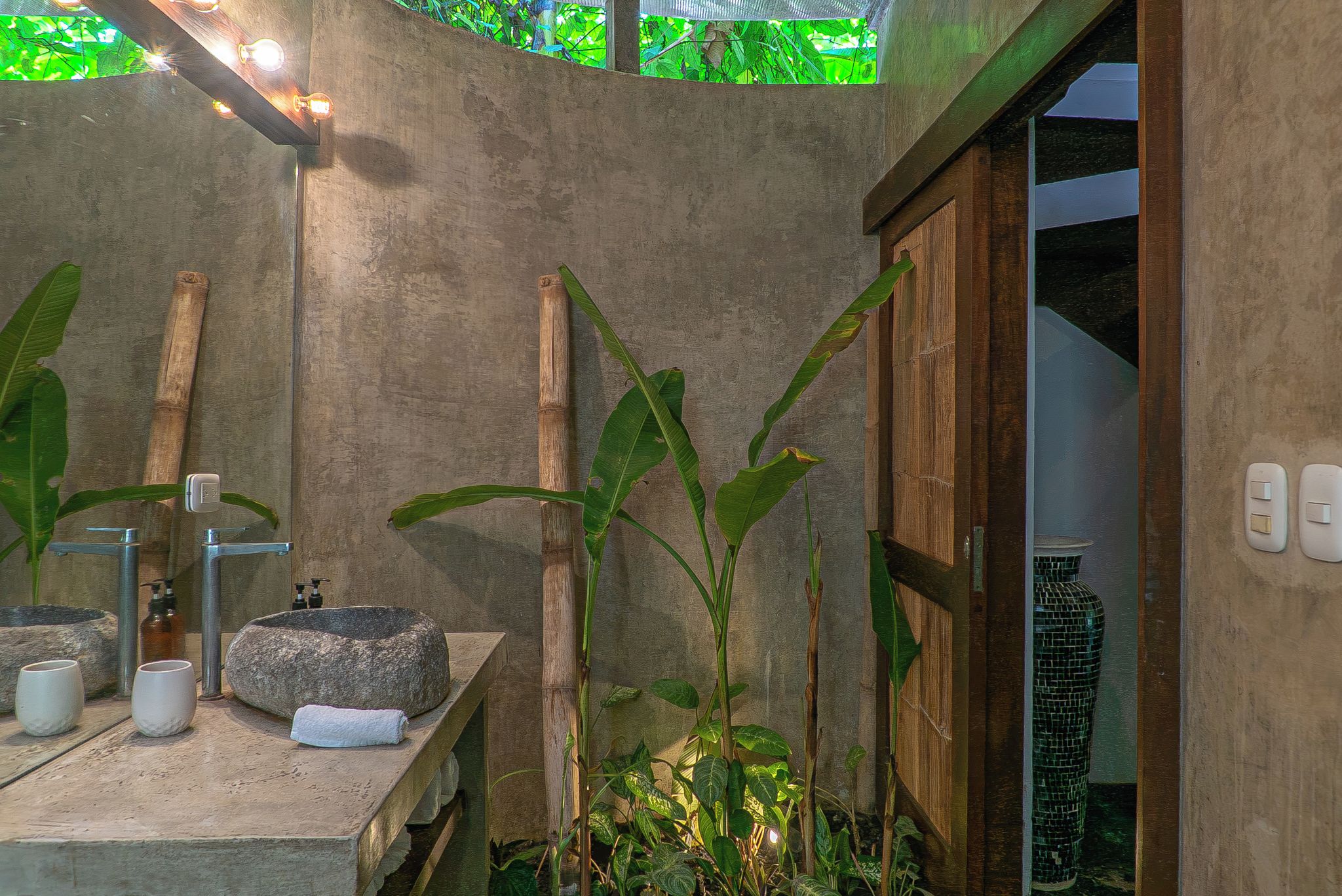 Casa de banho com parede de concreto, porta de madeira, lavatório com cuba de pedra e espelho.