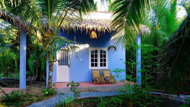 Uma casa azul com telhado de palha, rodeada por vegetação exuberante e palmeiras. Duas cadeiras de madeira estão na varanda.