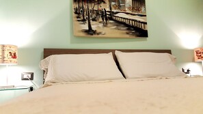 Cama com lençóis brancos, cabeceira de madeira e duas luminárias laterais com abajures estampados.