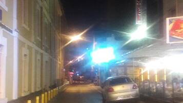 Cena noturna de rua com edifícios, um carro estacionado e luzes brilhantes.