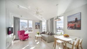 Uma sala de estar moderna com um sofá, uma poltrona rosa, uma mesa de jantar com cadeiras, um ventilador de teto e grandes janelas com vista para a cidade.