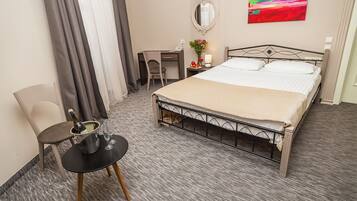 Quarto de hotel com cama, escrivaninha, cadeira, uma pequena mesa com uma garrafa de champanhe e um quadro colorido na parede.