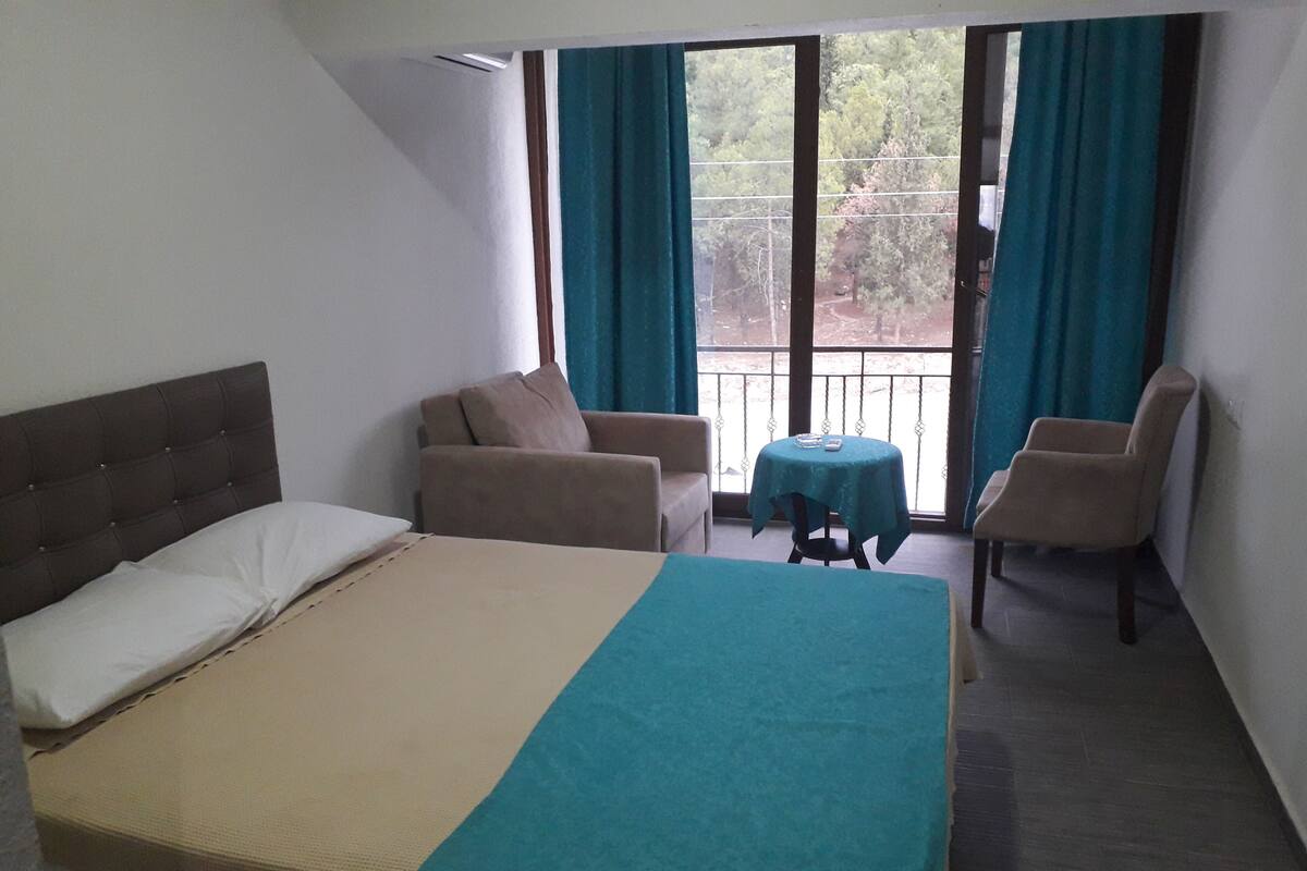 Quarto de hotel com cama, duas poltronas, uma mesinha e varanda com vista.