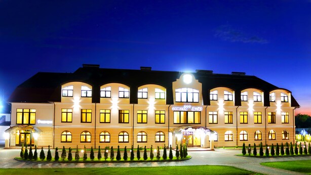 Um hotel grande, com vários andares, fachada simétrica, várias janelas e um gramado bem cuidado com sebes aparadas.