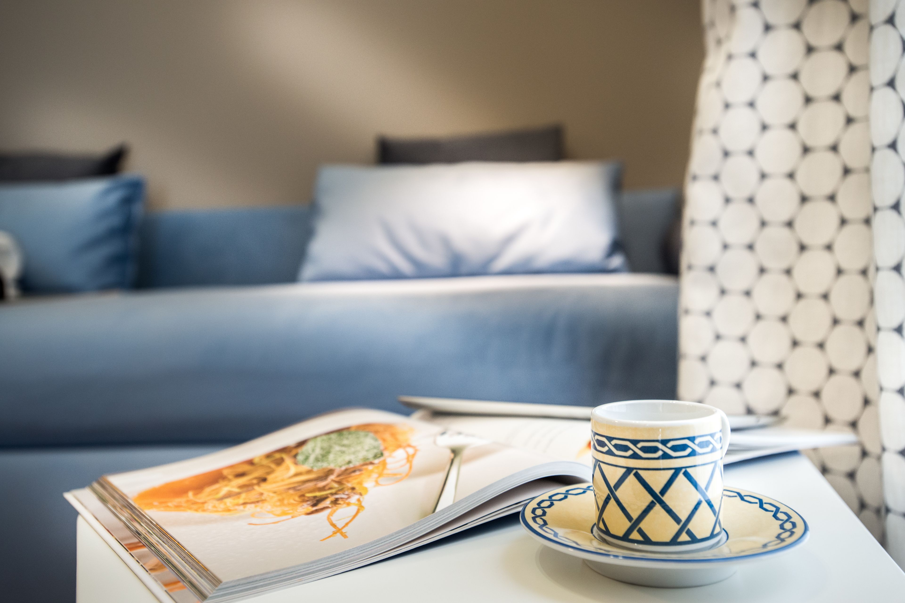 Uma cama com roupa de cama azul, cortina estampada e uma chávena sobre uma mesa.
