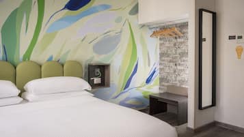 Quarto de hotel com cama, mesa de cabeceira, parede com arte abstrata, cabideiro e quadro emoldurado.