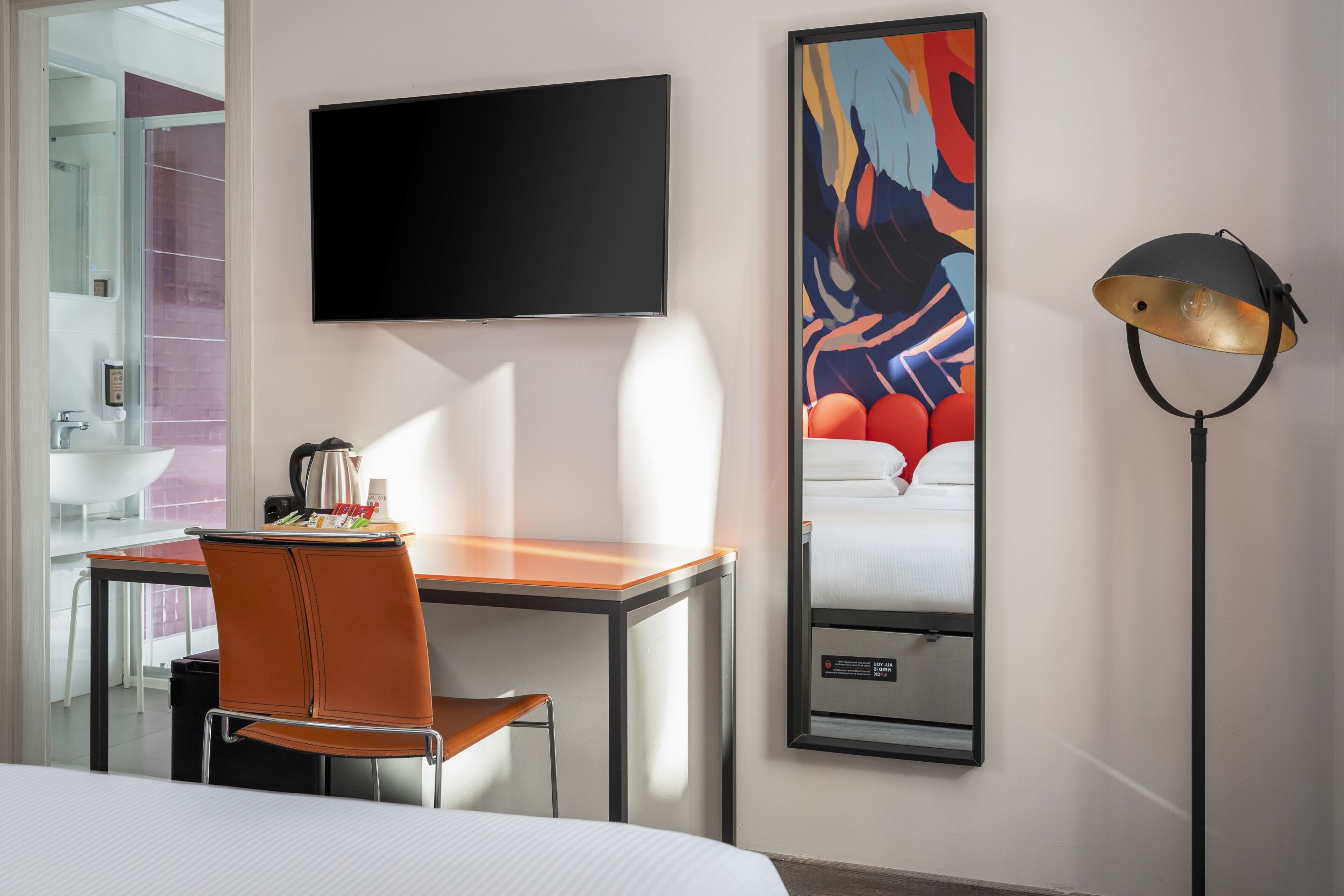 Quarto de hotel com uma cama, uma escrivaninha com cadeira, televisão, um quadro grande e um candeeiro de chão.