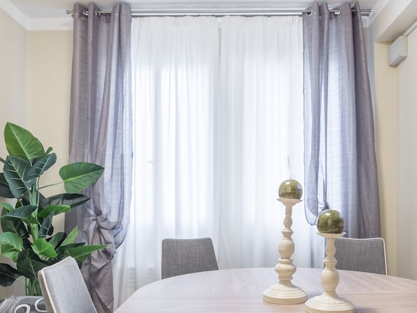 Uma área de refeições com uma mesa redonda, dois castiçais, um vaso de planta e cortinas cinzas.