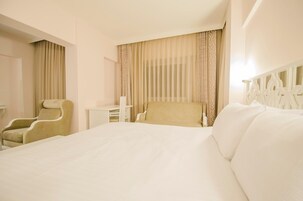 Quarto de hotel com uma cama grande, uma cadeira, uma escrivaninha e cortinas.