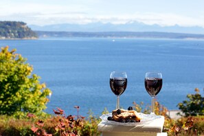 Vista panorâmica de uma área costeira com dois copos de vinho tinto sobre uma mesa, com vista para o mar e montanhas.