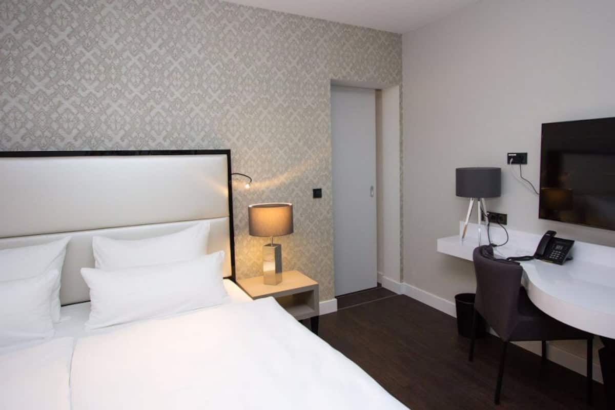 Quarto de hotel com cama, escrivaninha, cadeira, abajur, televisão e papel de parede estampado.