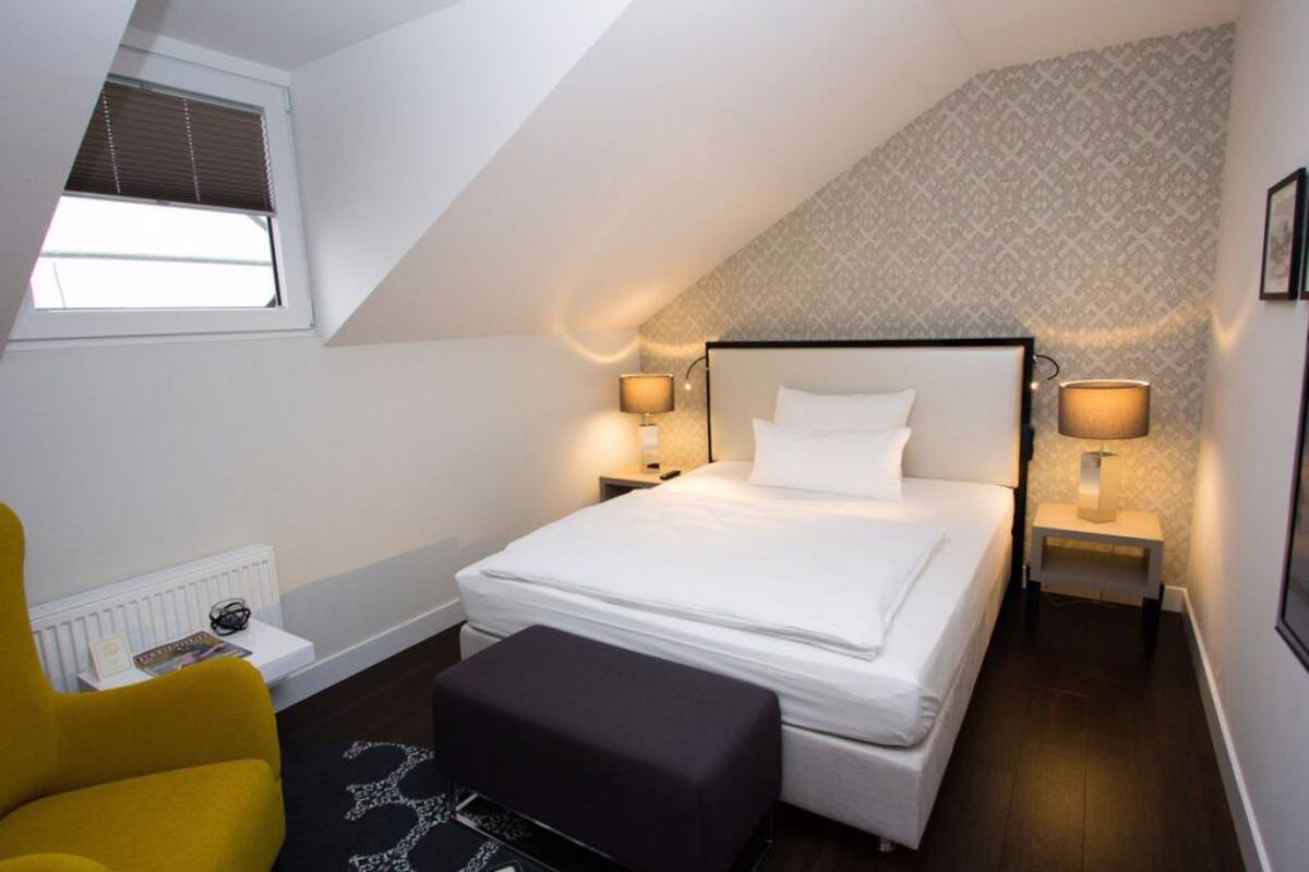 Quarto com cama, duas luminárias de cabeceira, janela com persianas, radiador, cadeira amarela e um banquinho preto.