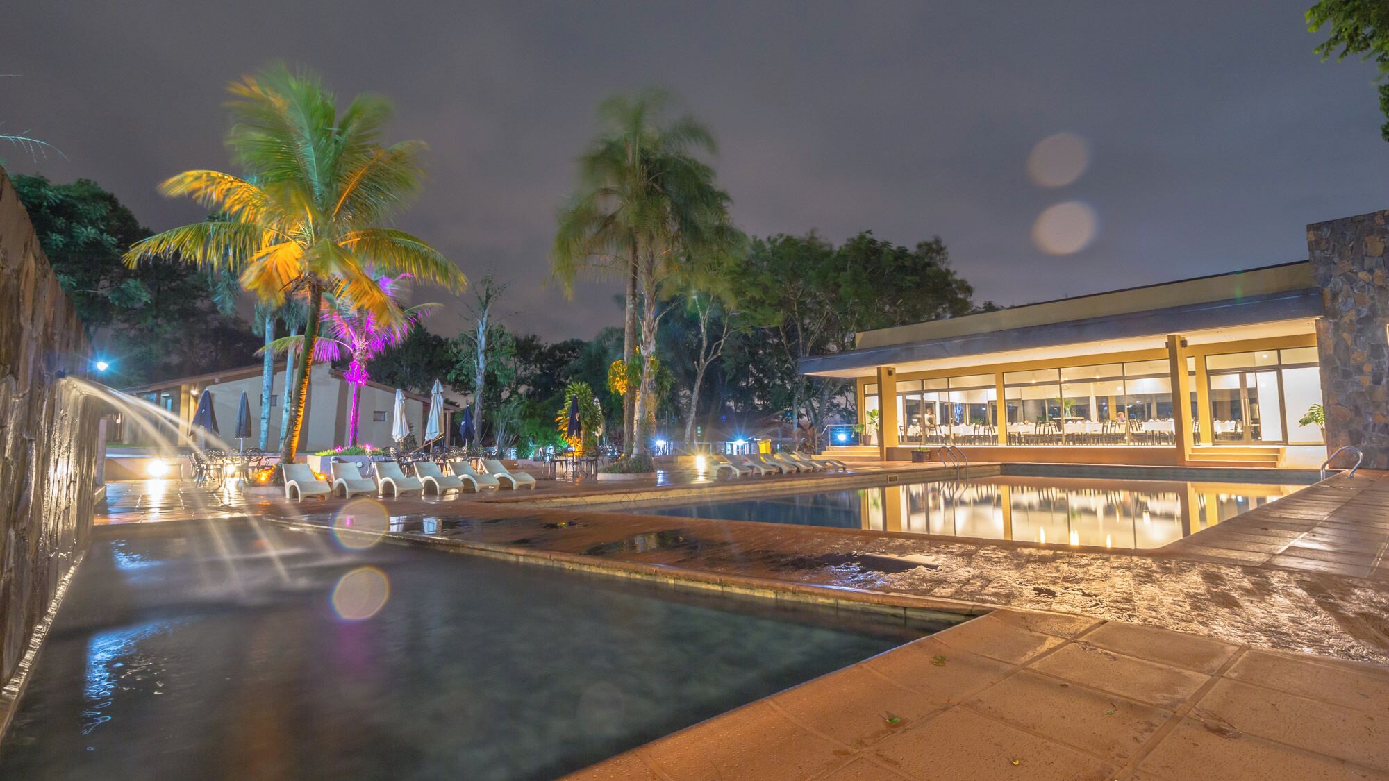 Uma piscina com espreguiçadeiras, palmeiras e um edifício com grandes janelas.