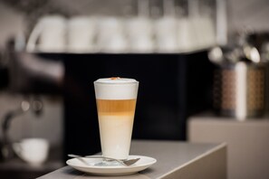Um copo alto com bebida em camadas sobre um prato branco e uma colher, com o fundo desfocado de um bar.
