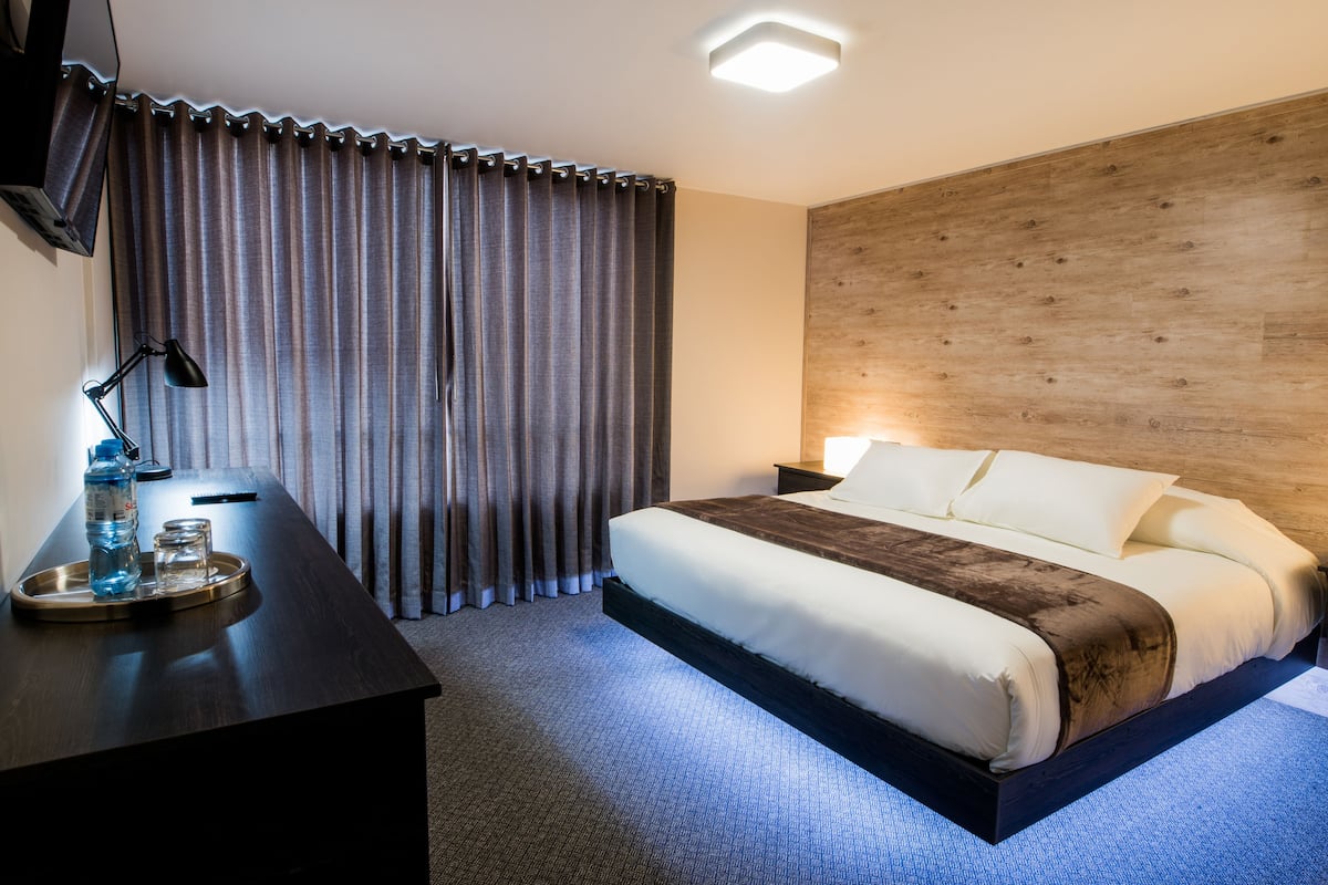 Quarto de hotel com cama, escrivaninha, luminária, garrafa de água e mesa de cabeceira.