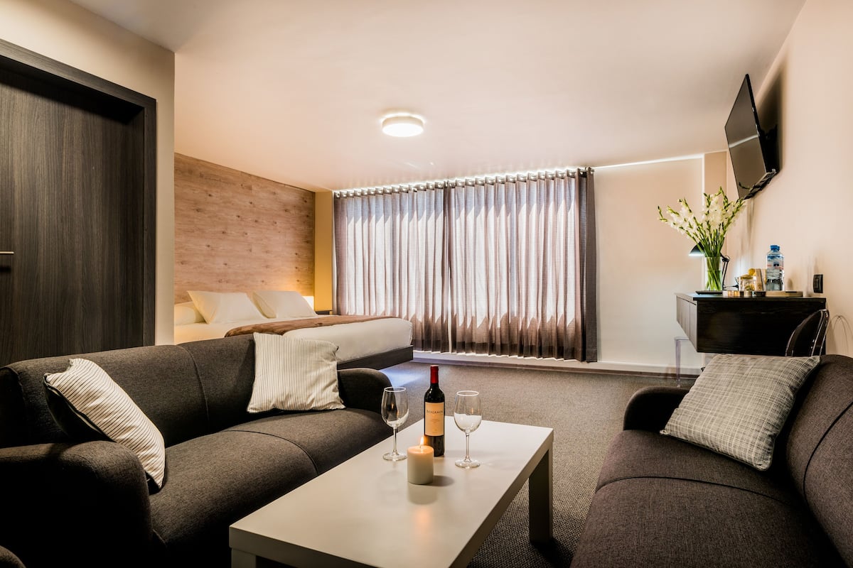 Uma sala de estar moderna com um sofá, uma mesa de centro com uma vela e taças de vinho, uma cama com cortinas e uma TV fixada na parede.