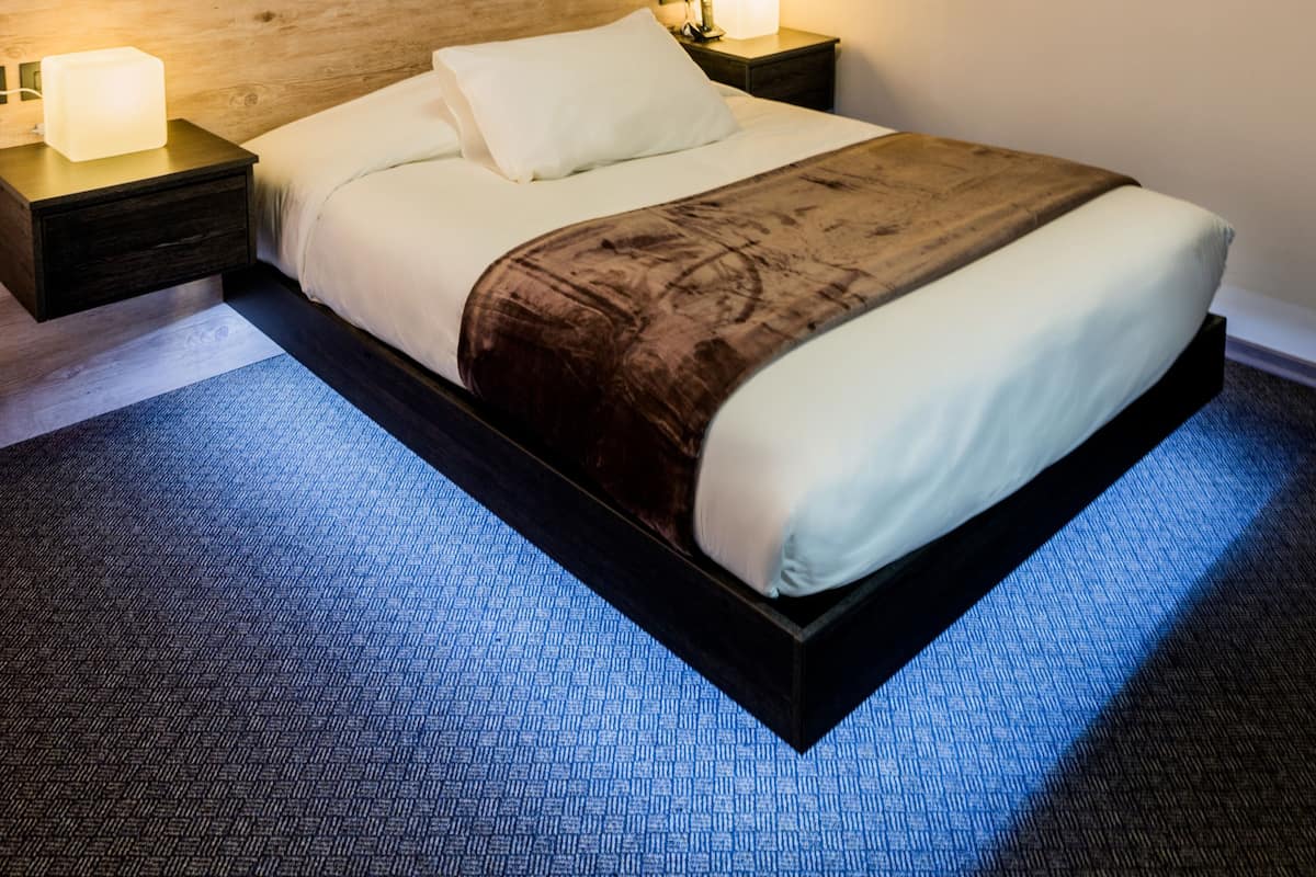 Quarto de hotel com cabeceira de madeira, cama com roupa de cama branca, duas luminárias laterais e um tapete azul.