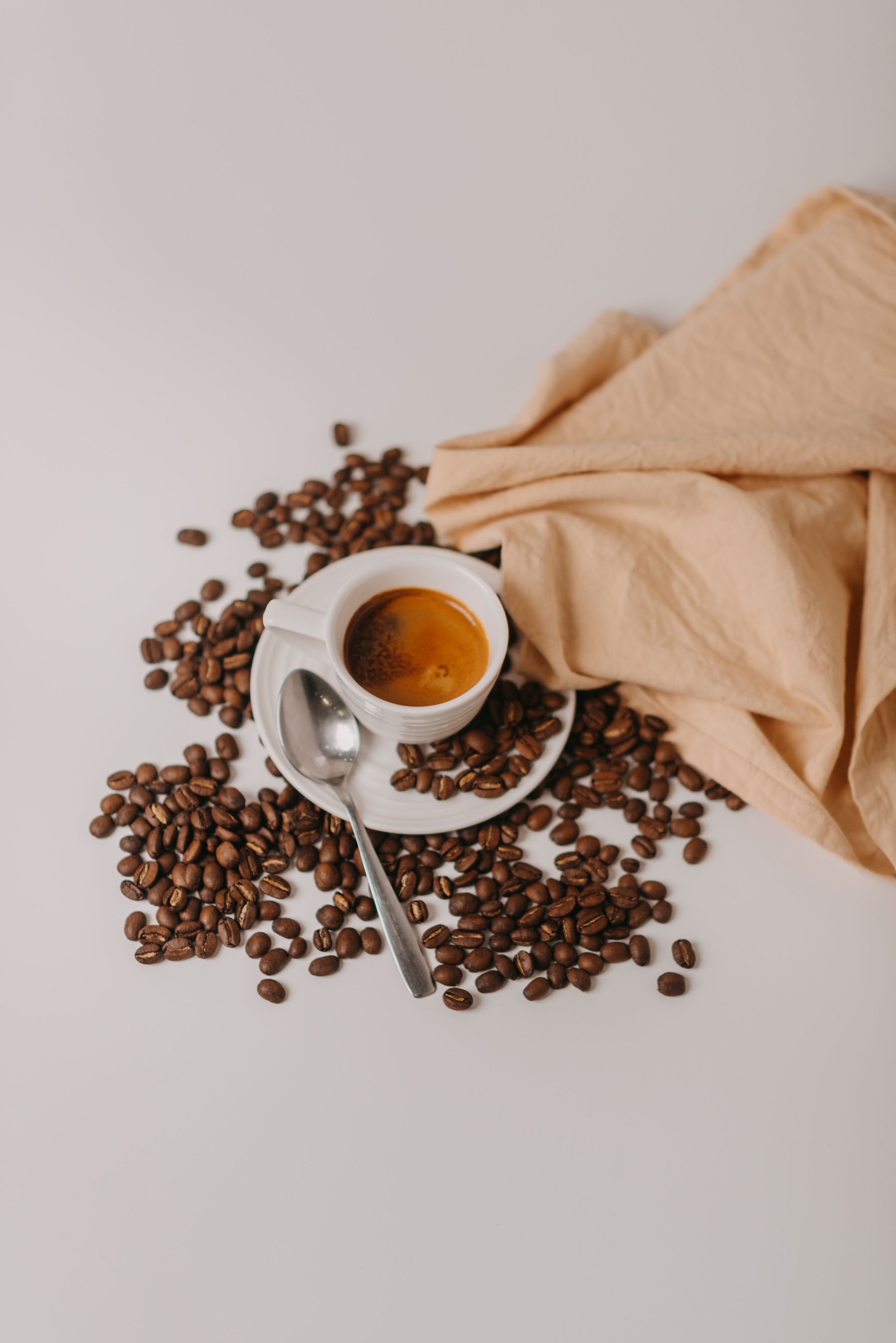 Uma chávena de café com uma colher num pires, rodeada por grãos de café sobre uma superfície branca.