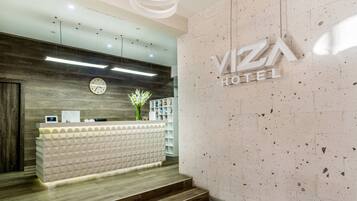 Uma receção com um relógio, uma parede com o letreiro "VIZA HOTEL" e uma escadaria que leva ao andar superior.