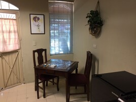 Uma área de refeições com uma mesa, cadeiras, uma janela com persianas, uma planta pendente e um eletrodoméstico preto.