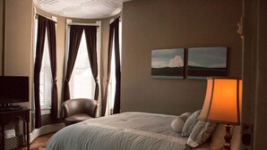Quarto de hotel com uma cama, uma cadeira, uma TV e duas pinturas na parede.