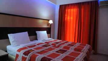 Quarto de hotel com cama, mesa de cabeceira, luminária e janela com cortinas vermelhas.