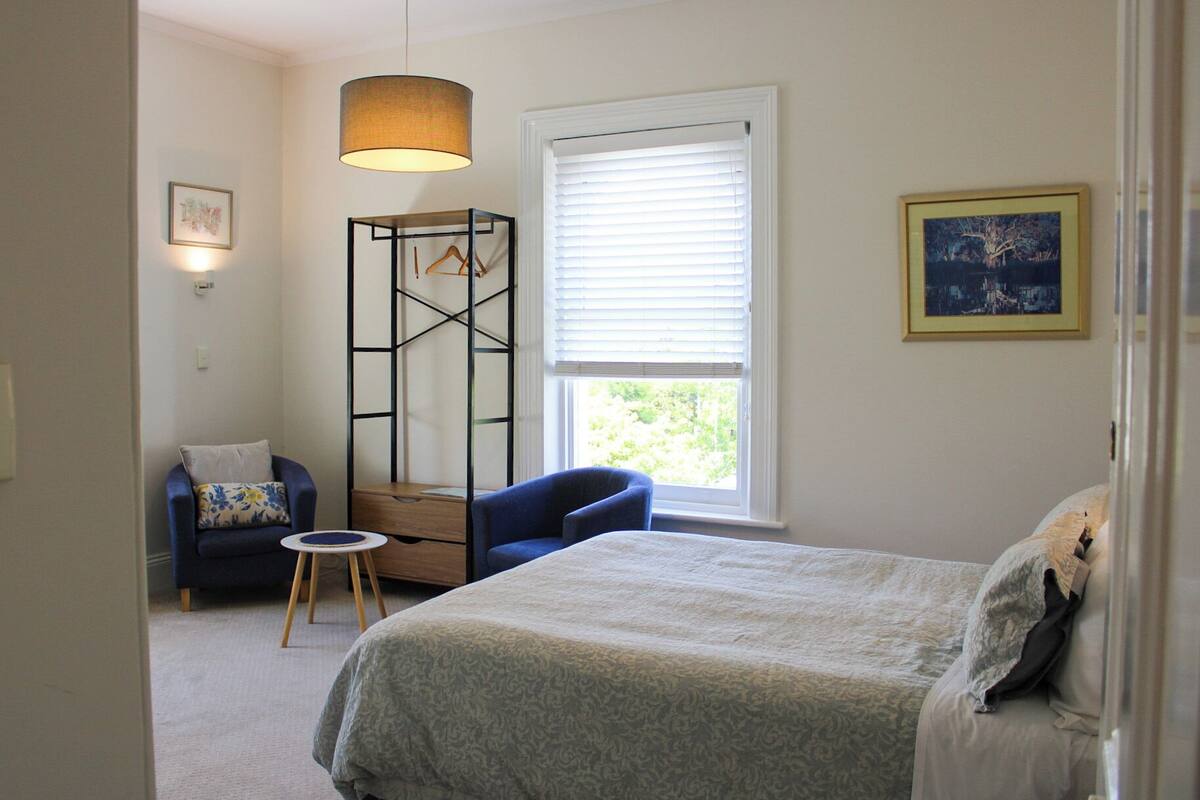 Quarto com cama, cadeira, luminária pendente, janela com persianas e quadro na parede.