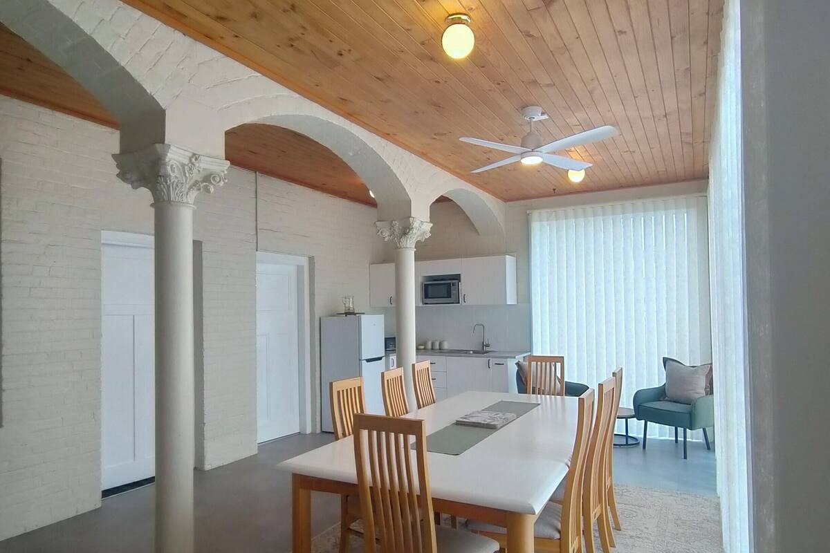 Uma sala de jantar espaçosa com uma mesa comprida e cadeiras, cozinha ao fundo e um ventilador de teto.