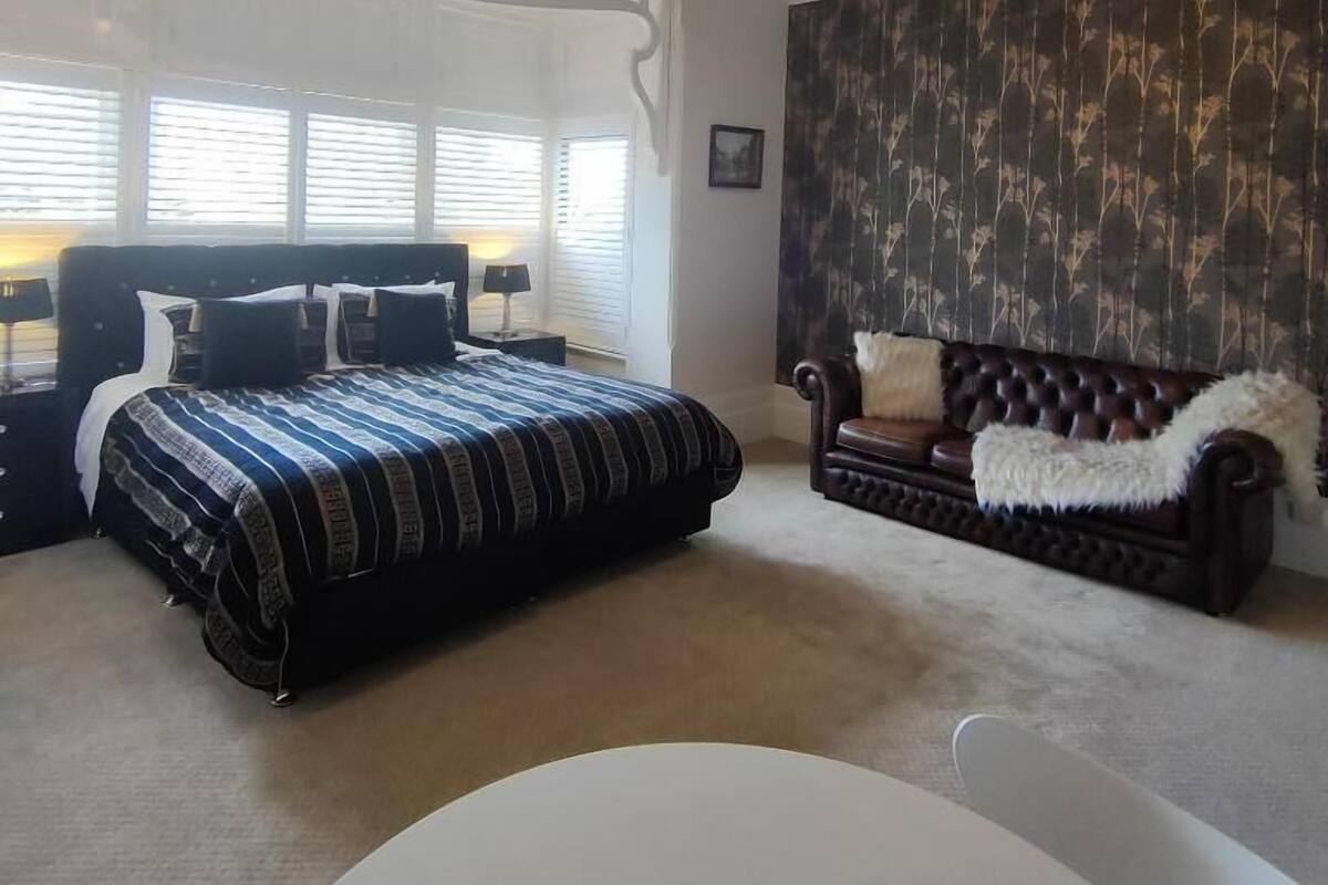 Quarto com uma cama grande, um sofá marrom escuro com manta de pele branca e papel de parede estampado.