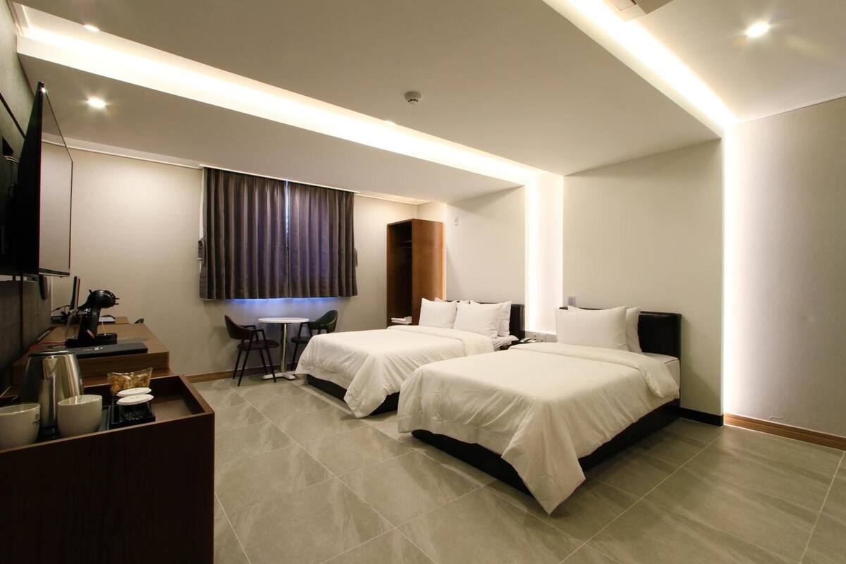 Quarto de hotel moderno com duas camas, uma escrivaninha com cadeira, televisão e uma pequena mesa com chávenas e chaleira.