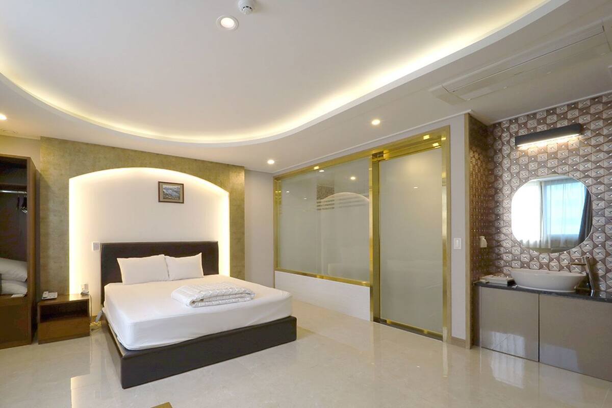 Quarto moderno com cama, mesas de cabeceira, guarda-roupa, banheiro com pia e espelho, e divisória de vidro.