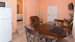 Quarto compacto com uma mesa redonda de madeira, duas cadeiras, uma geladeira branca e uma porta que dá para outro cômodo.