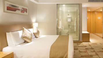 Quarto de hotel com uma cama grande, uma mesa de cabeceira, um abajur e um box de chuveiro de vidro.