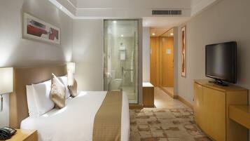 Quarto de hotel com uma cama grande, mesas de cabeceira, uma televisão e um banheiro visível através de uma porta de vidro.