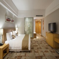 Quarto de hotel com uma cama grande, mesas de cabeceira, uma televisão e um banheiro visível através de uma porta de vidro.