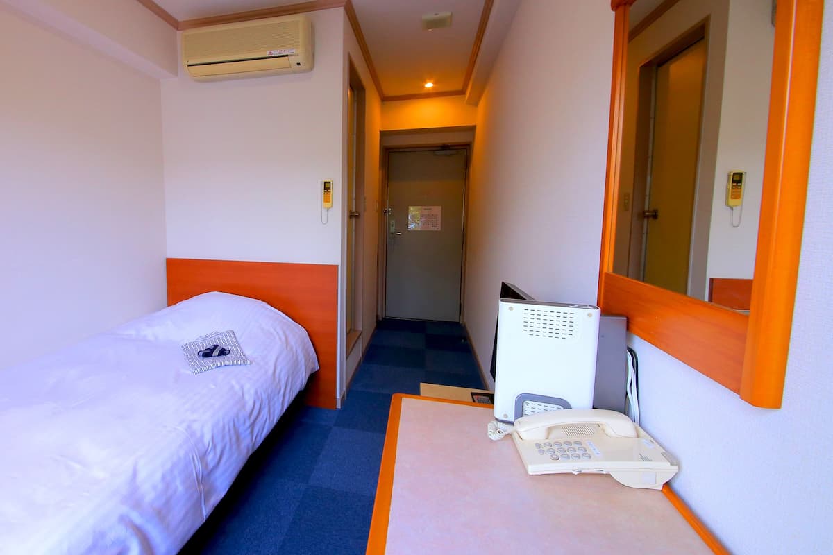 Quarto de hotel estreito com uma cama de solteiro, uma escrivaninha com telefone e um computador.