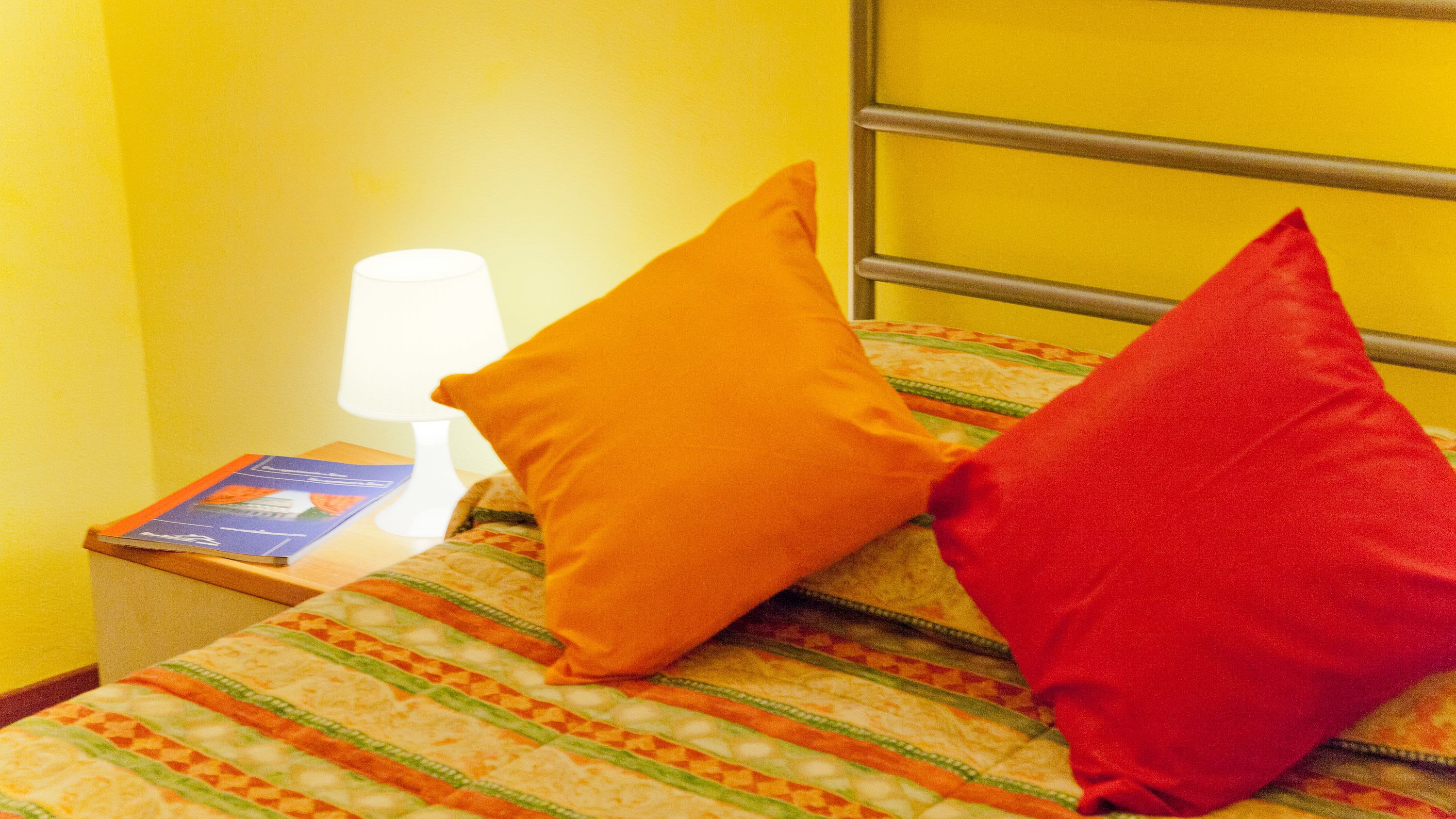 Cama com duas almofadas, mesa de cabeceira com um livro e uma luminária, e parede amarela.