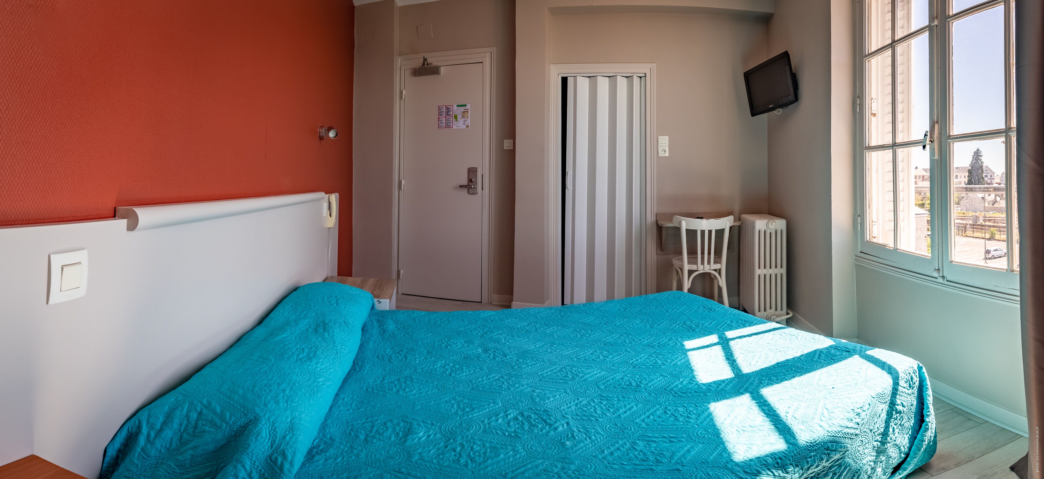 Quarto com cama, janela, radiador, porta e televisão na parede.