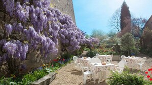Uma área externa com mesas e cadeiras, rodeada por um jardim com flores roxas e um muro de pedra.