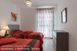 Quarto com duas camas, uma mesa de cabeceira, luminária, cômoda e janela com cortinas florais.