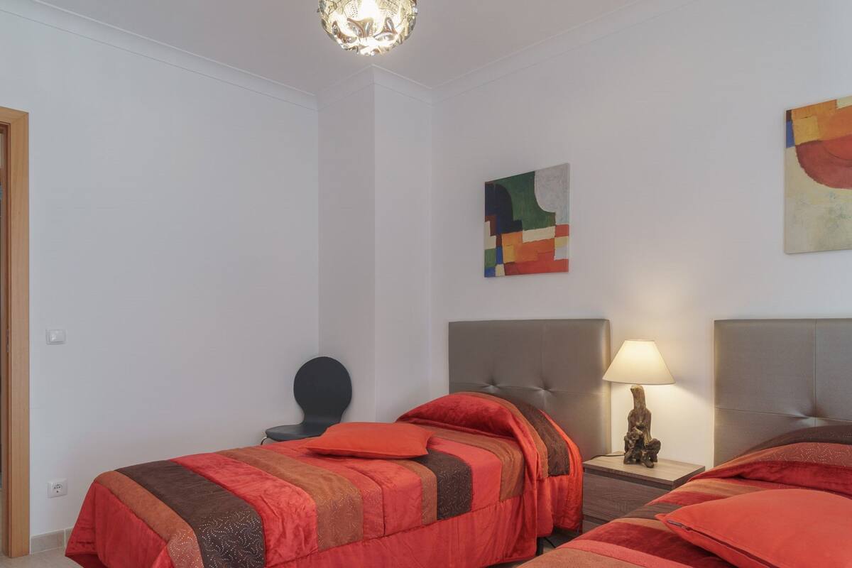 Quarto de hotel com duas camas, uma mesa de cabeceira com luminária, um quadro na parede e um banheiro visível por uma porta aberta.