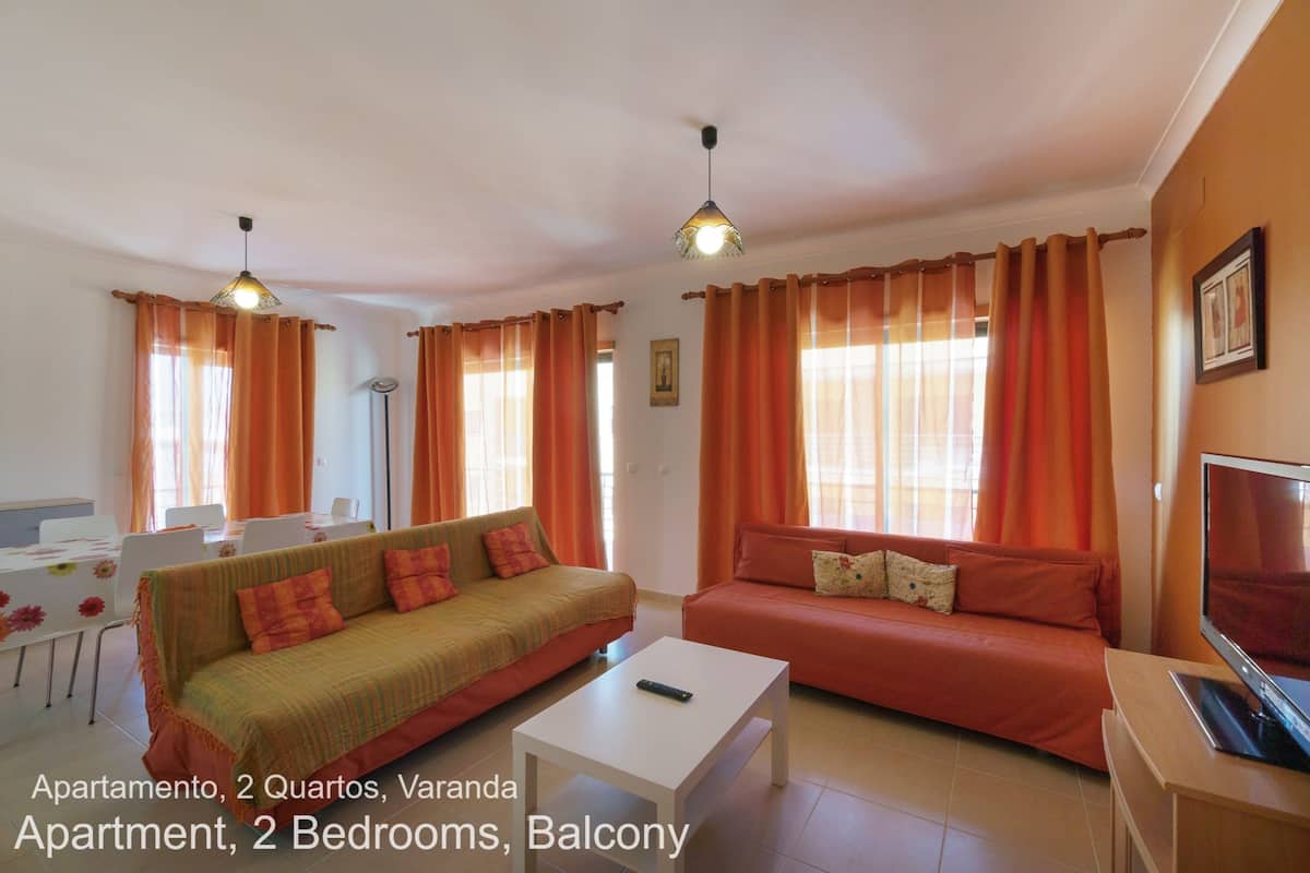 Apartamento de 2 quartos com varanda, com dois sofás, uma mesa de jantar e uma televisão.