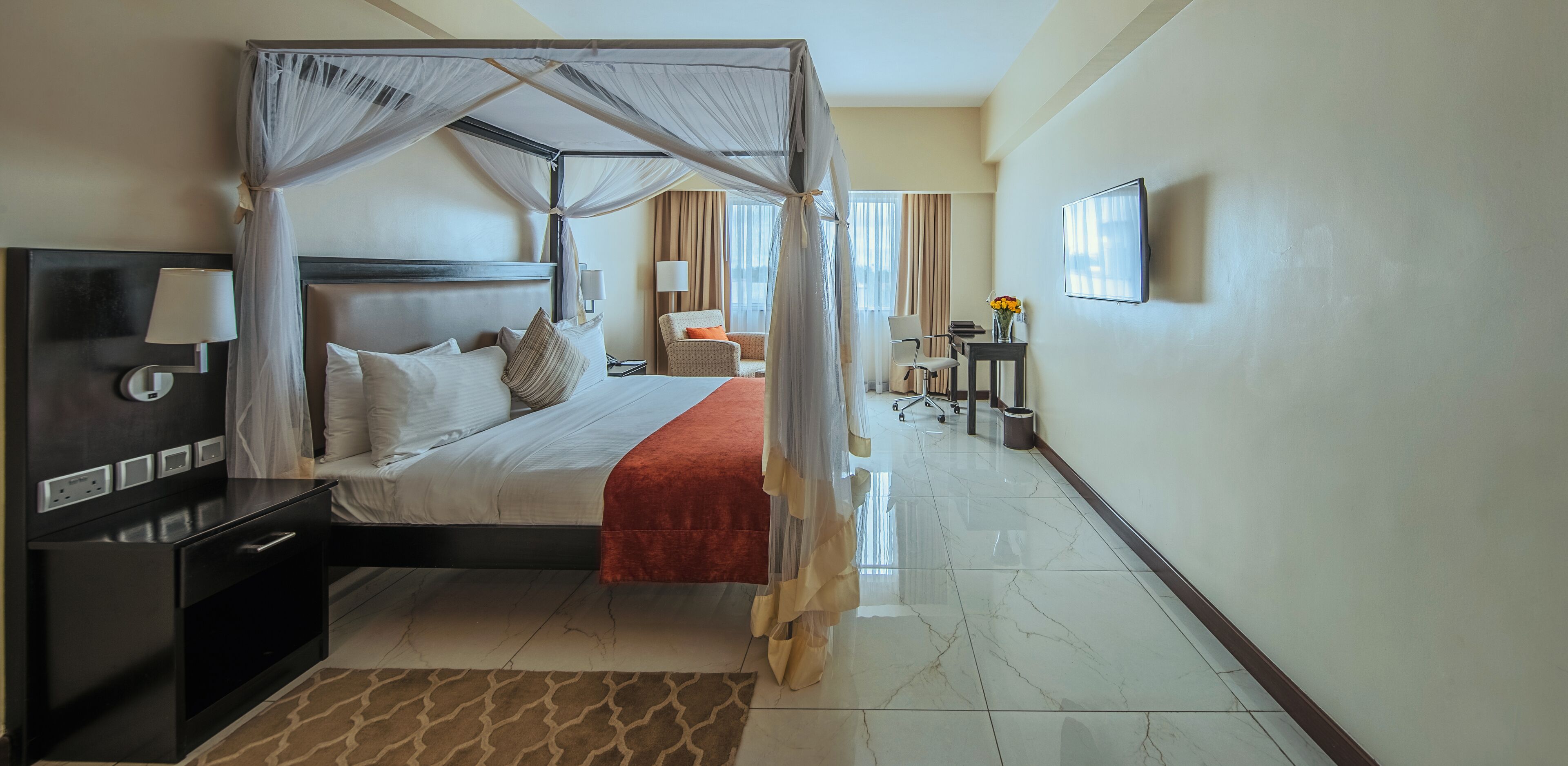 Quarto de hotel com cama de dossel, escrivaninha com luminária, uma cadeira e uma pequena mesa com um vaso de flores.