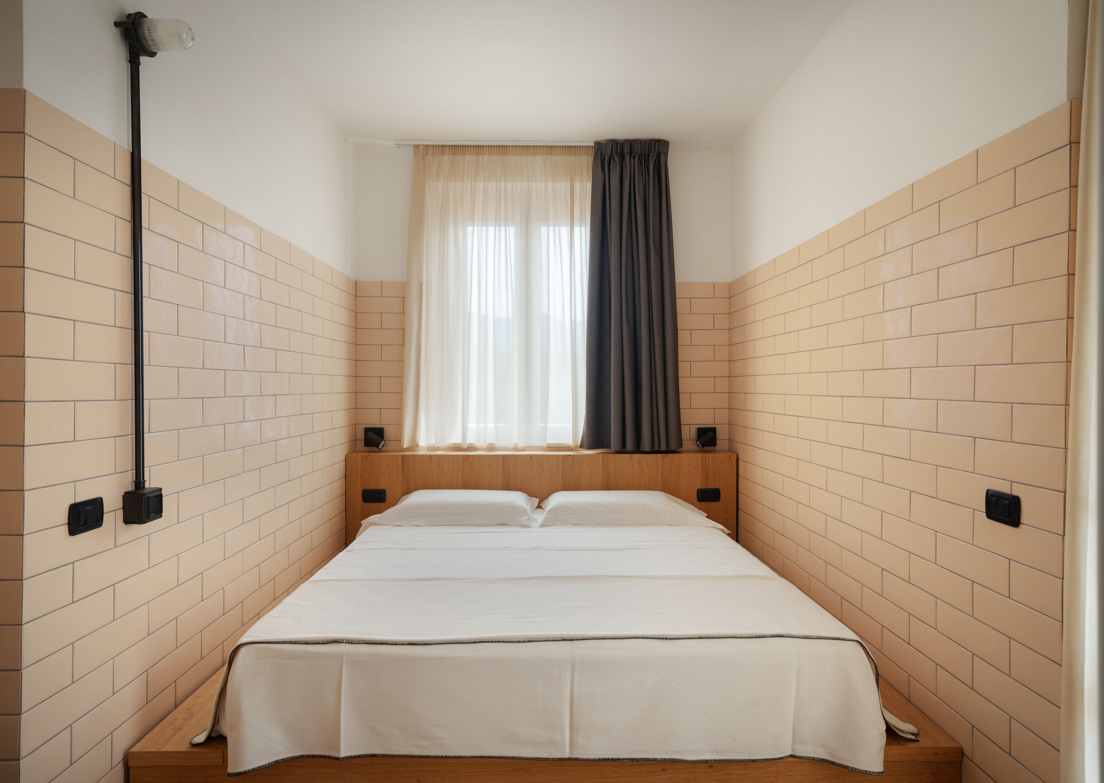 Quarto de hotel estreito com uma cama de solteiro, janela com cortinas finas e luminária fixada na parede.