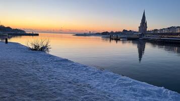 Vista para a orla marítima com um caminho coberto de neve, uma torre alta e água calma refletindo o céu.