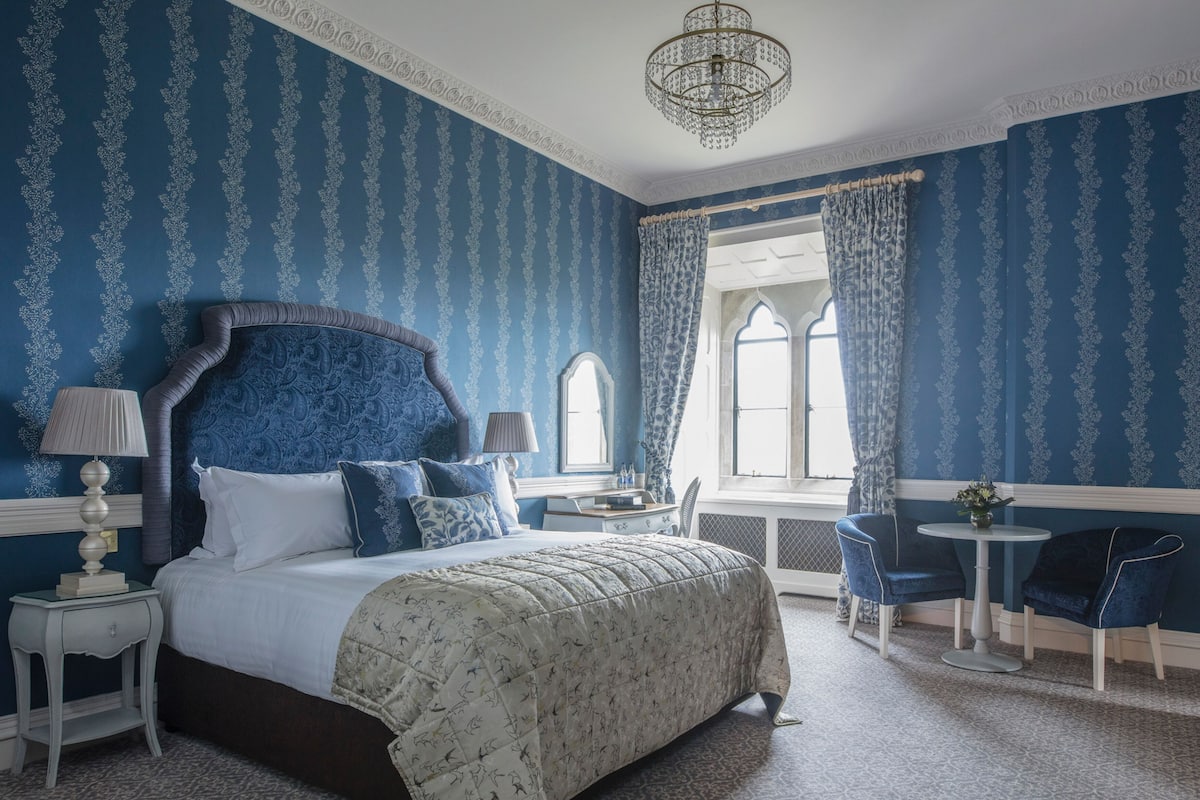 Quarto com cama grande, escrivaninha e cadeira. Tem uma janela com cortinas e um candelabro.