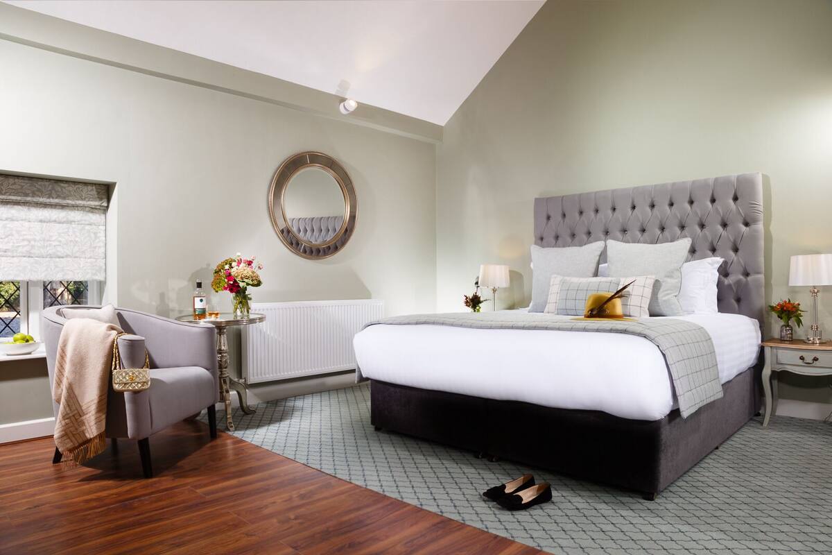 Quarto moderno com uma cama grande, um espelho redondo, uma pequena mesa com um vaso de flores e uma cadeira com uma bolsa.