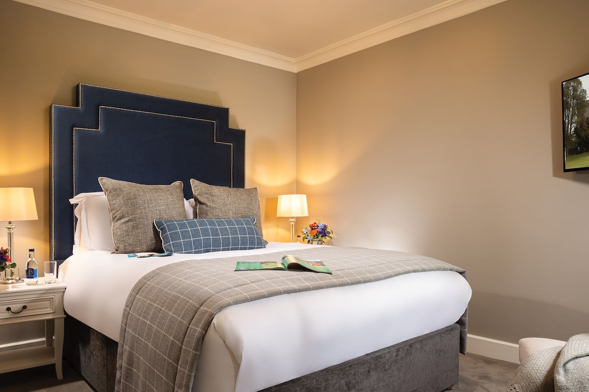Quarto de hotel com uma cama grande, duas mesas de cabeceira, um criado-mudo, uma luminária, uma revista e um quadro na parede.